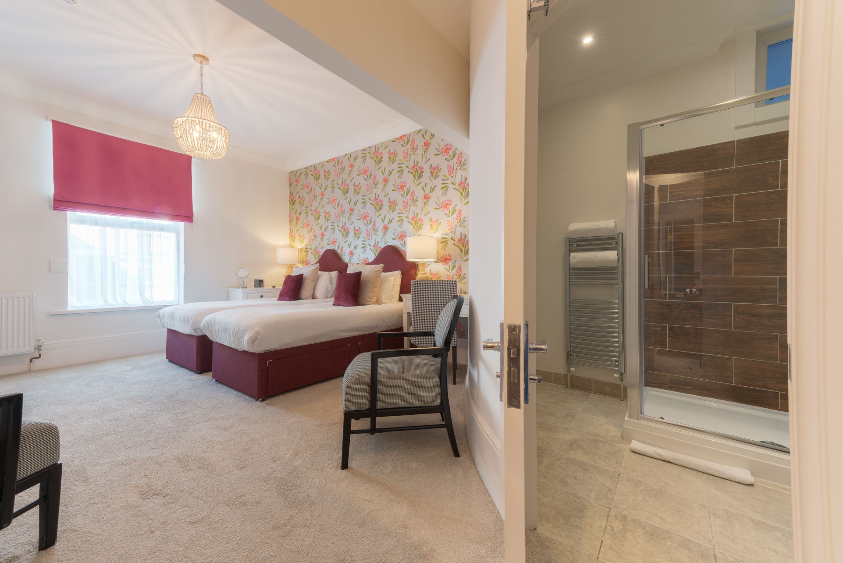 Quarto com cama, mesas de cabeceira, uma cadeira, janela com cortinas, área de chuveiro com porta de vidro e parede estampada.