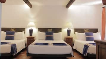 Quarto de hotel com uma cama grande, duas camas menores e uma mesinha de cabeceira com luminárias. O quarto possui piso em azulejo e teto de madeira.