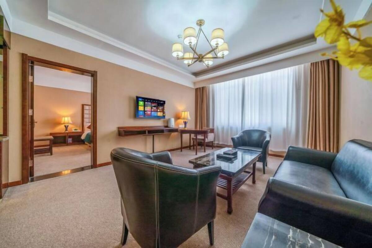 Quarto de hotel com área de estar, televisão e uma janela grande com cortinas.