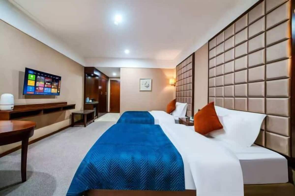 Quarto de hotel com cama, escrivaninha, televisão e luminária fixada na parede.