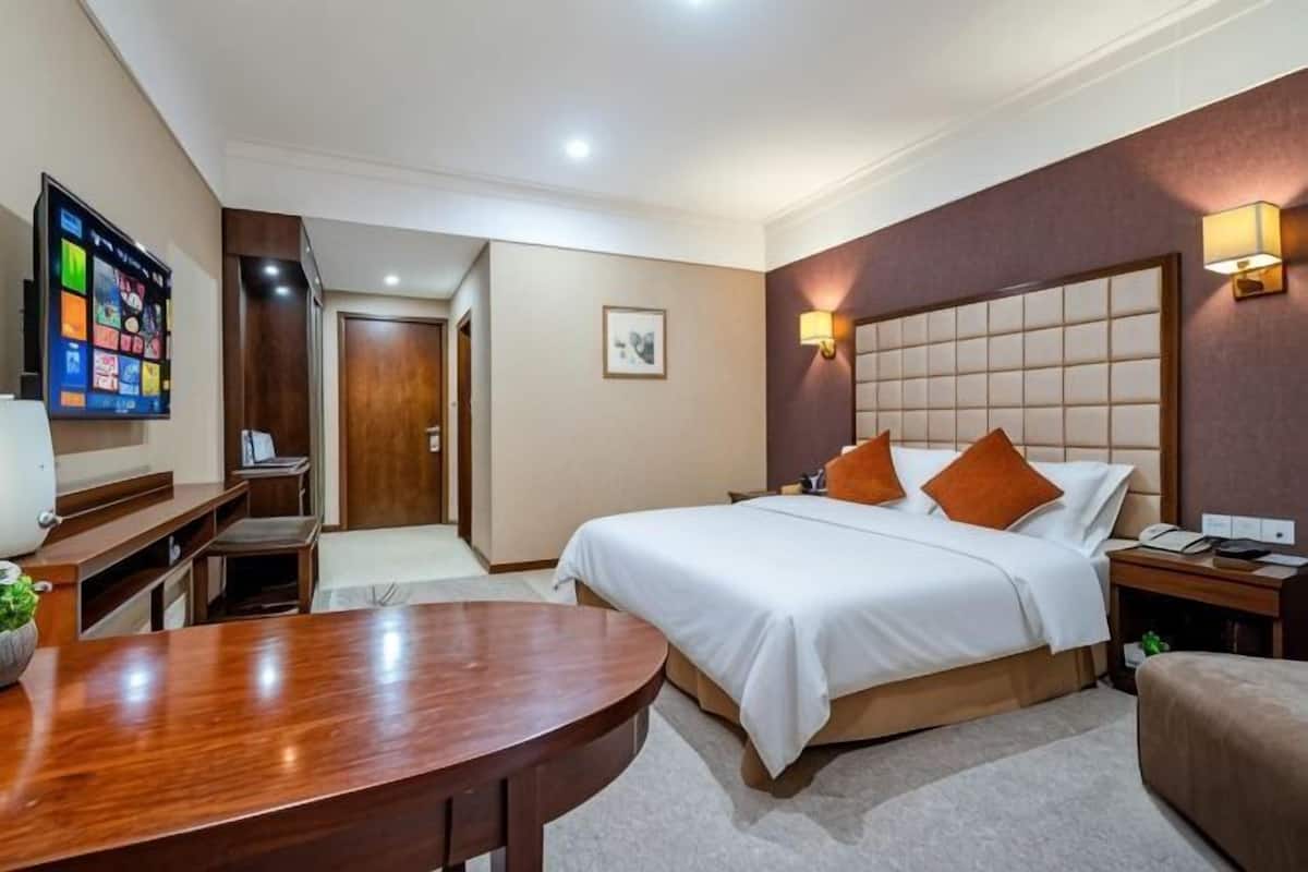 Quarto de hotel com cama, mesa de cabeceira, escrivaninha, cadeira e televisão na parede.