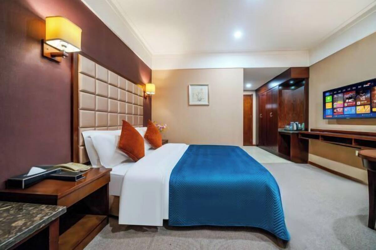 Quarto de hotel com cama, mesa de cabeceira, luminária, televisão e guarda-roupa.