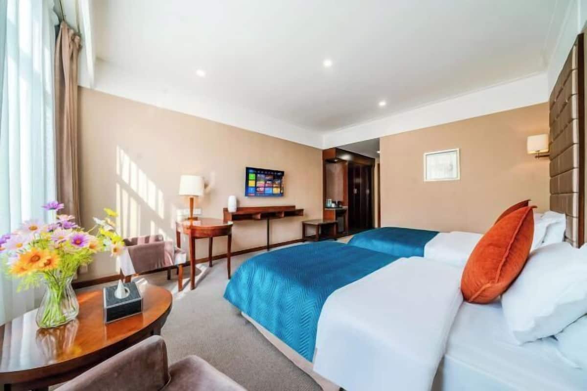 Quarto de hotel com cama, escrivaninha, cadeira, mesinha com flores e janela com cortinas.