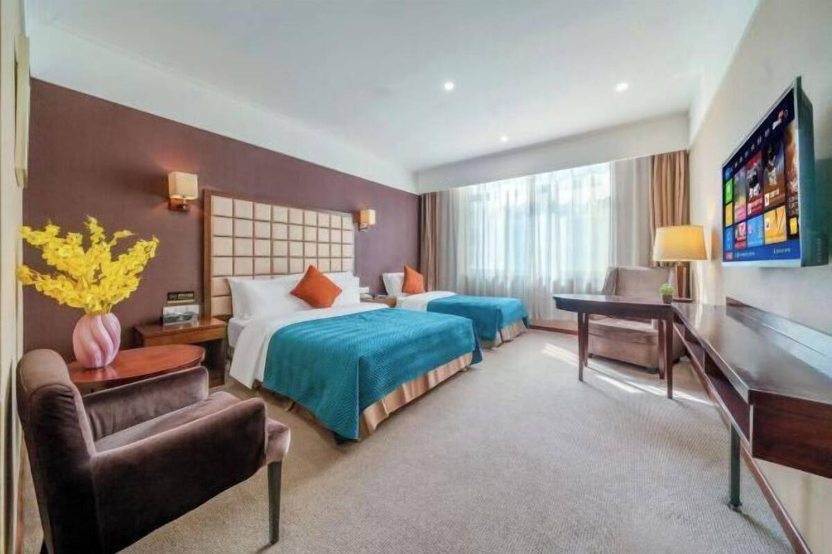 Quarto de hotel com duas camas, uma escrivaninha, uma cadeira, televisão e um vaso com flores.