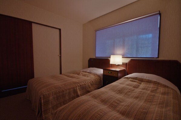 Quarto de hotel com duas camas de solteiro, uma mesa de cabeceira com luminária, tela de projeção e uma porta.