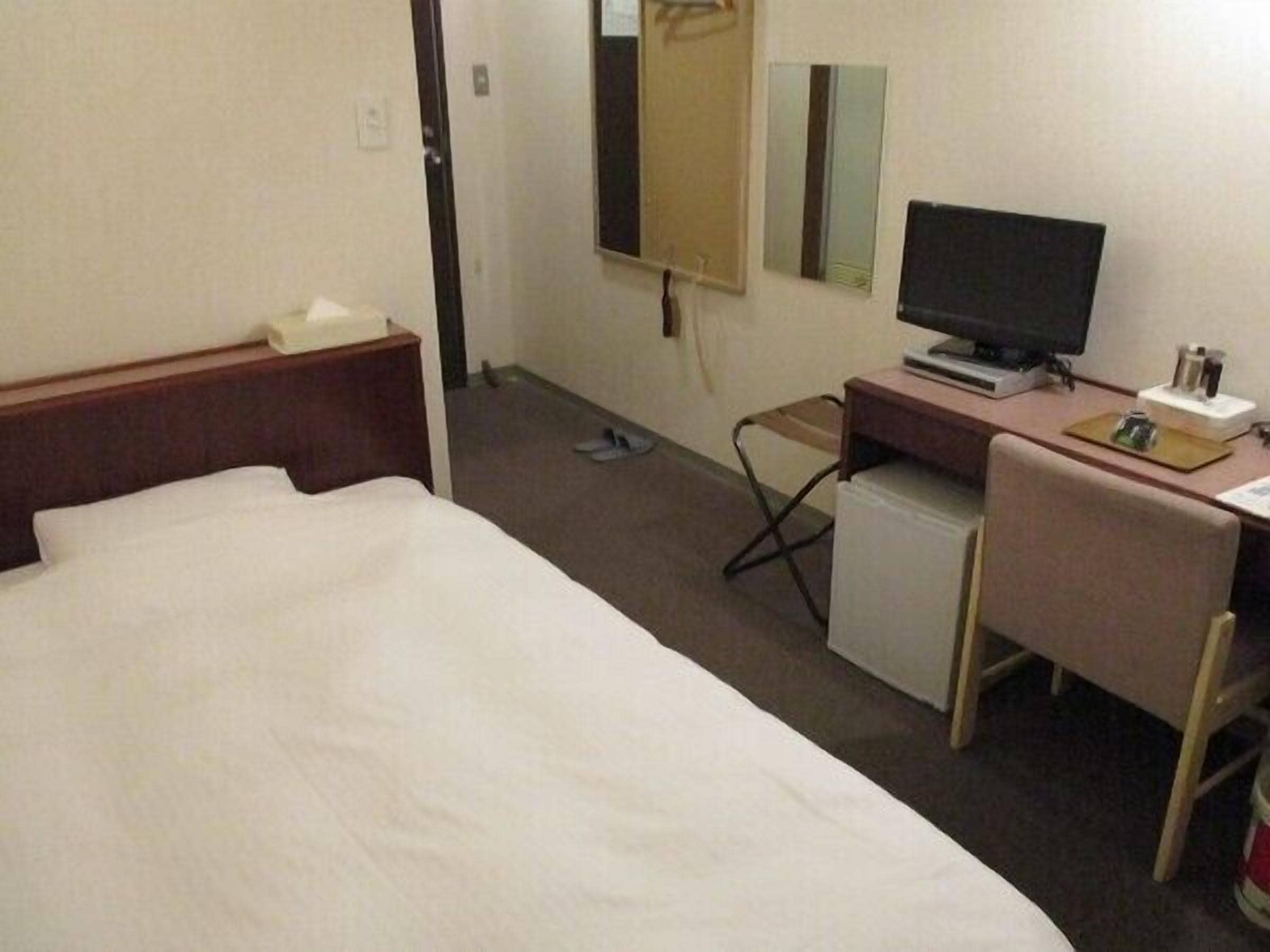Quarto de hotel com cama, escrivaninha com computador, cadeira e um pequeno frigorífico.