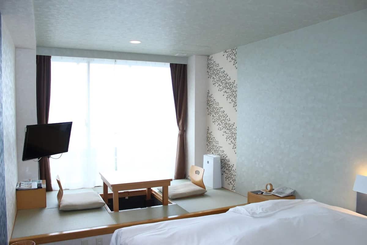 Quarto de hotel com cama, escrivaninha, cadeira, televisão, mesa de cabeceira e parede com padrão de folhas.
