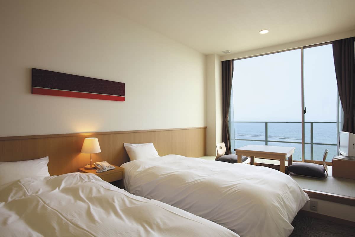 Quarto de hotel com duas camas, uma mesa, uma cadeira, um abajur e uma janela com vista para o mar.