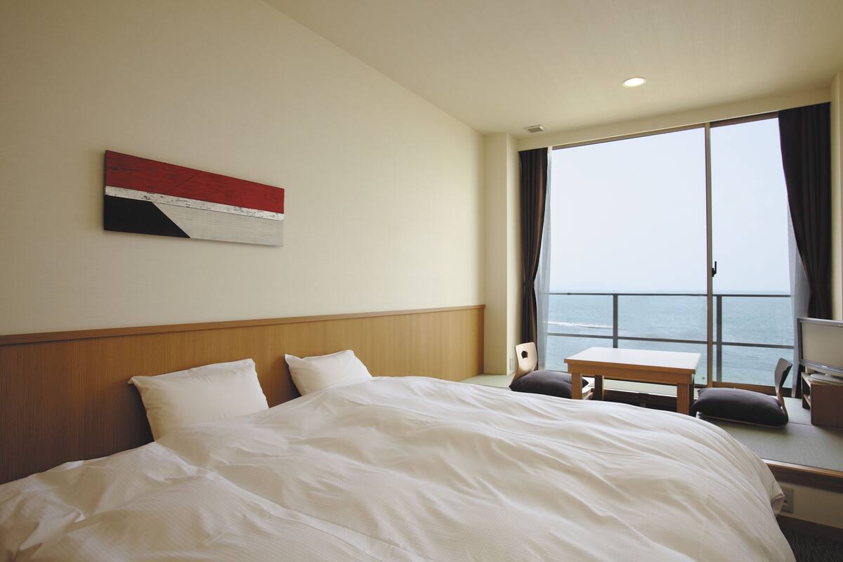 Quarto de hotel com uma cama grande, uma mesinha, uma cadeira e vista para o mar através de uma janela ampla.