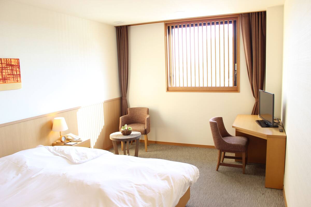 Quarto de hotel com cama, escrivaninha, cadeira, mesa pequena e janela com cortinas.