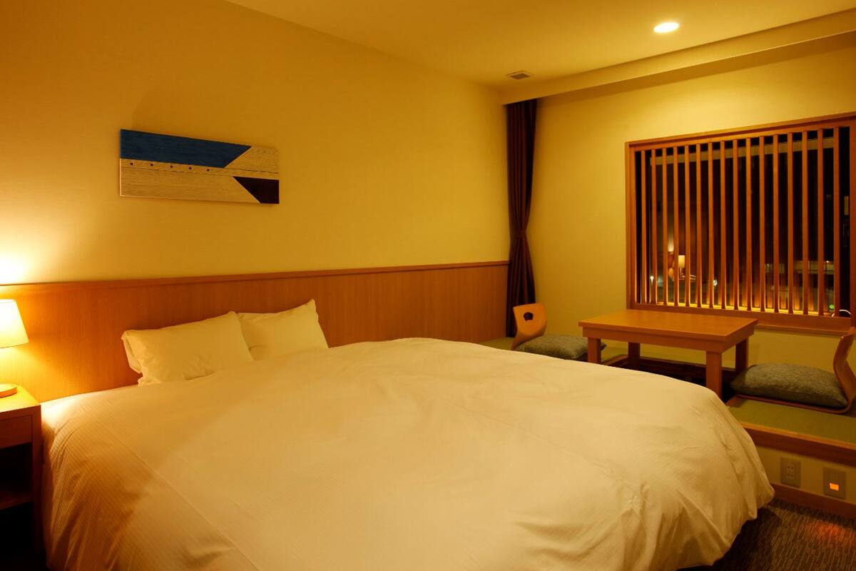 Quarto de hotel com uma cama grande, uma escrivaninha, uma cadeira, um abajur e uma janela com persianas.