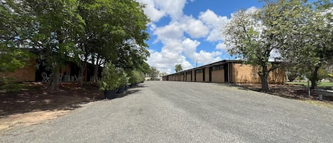 Uma estrada de cascalho que leva a uma fileira de edifícios de tijolos com grandes janelas, cercada por árvores.