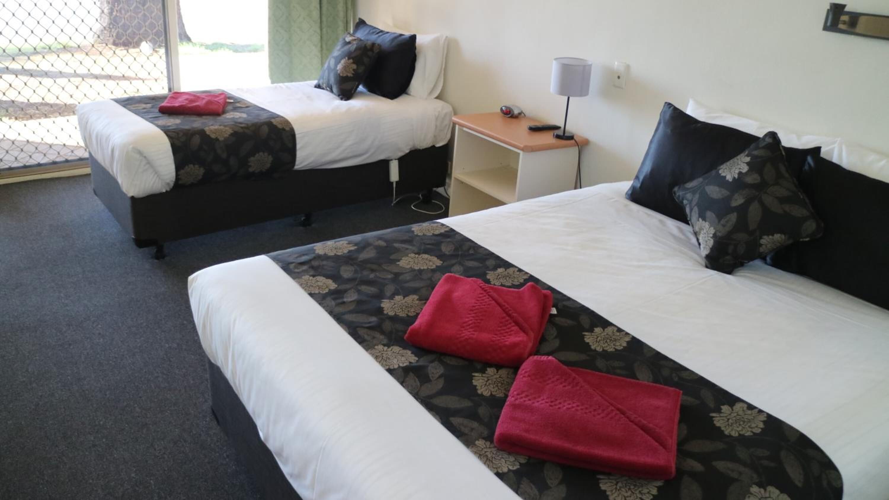 Quarto de hotel com duas camas, cada uma com colcha e almofadas florais, uma pequena mesa de cabeceira de madeira com abajur, e vista pela janela com grade de arame.