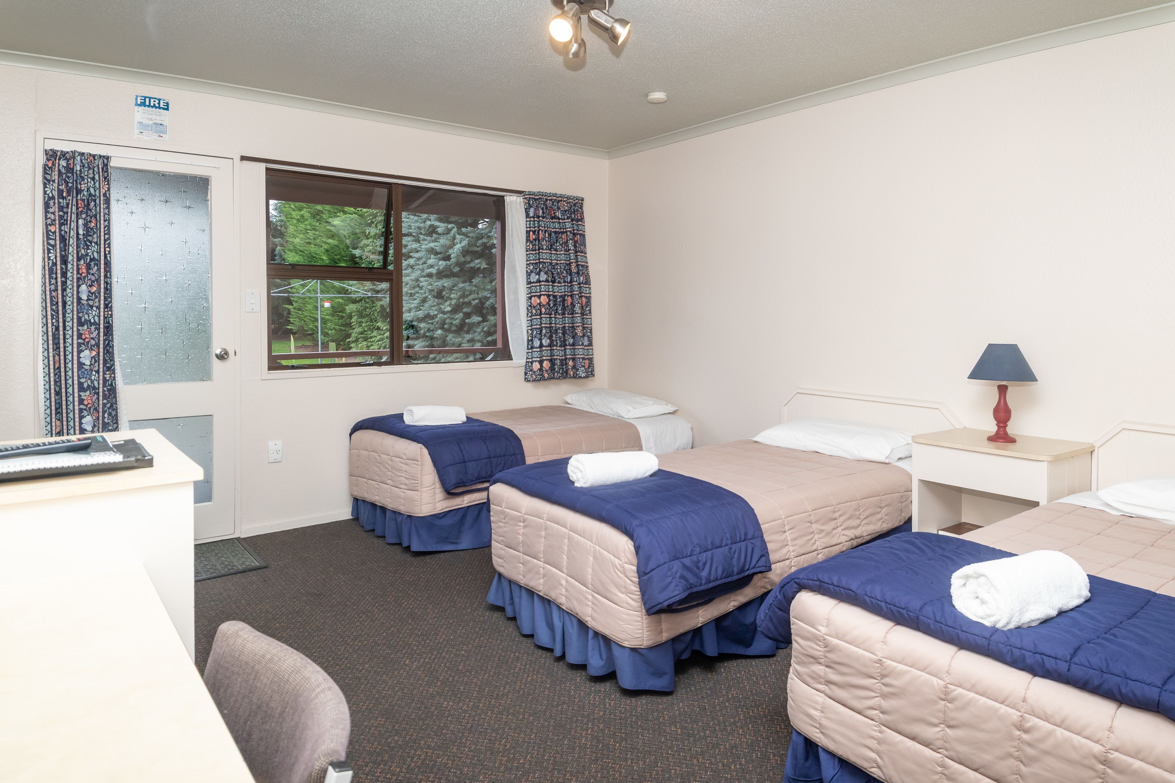 Quarto de hotel com três camas, uma escrivaninha, uma cadeira, uma janela com cortinas e uma mesa de cabeceira com abajur.