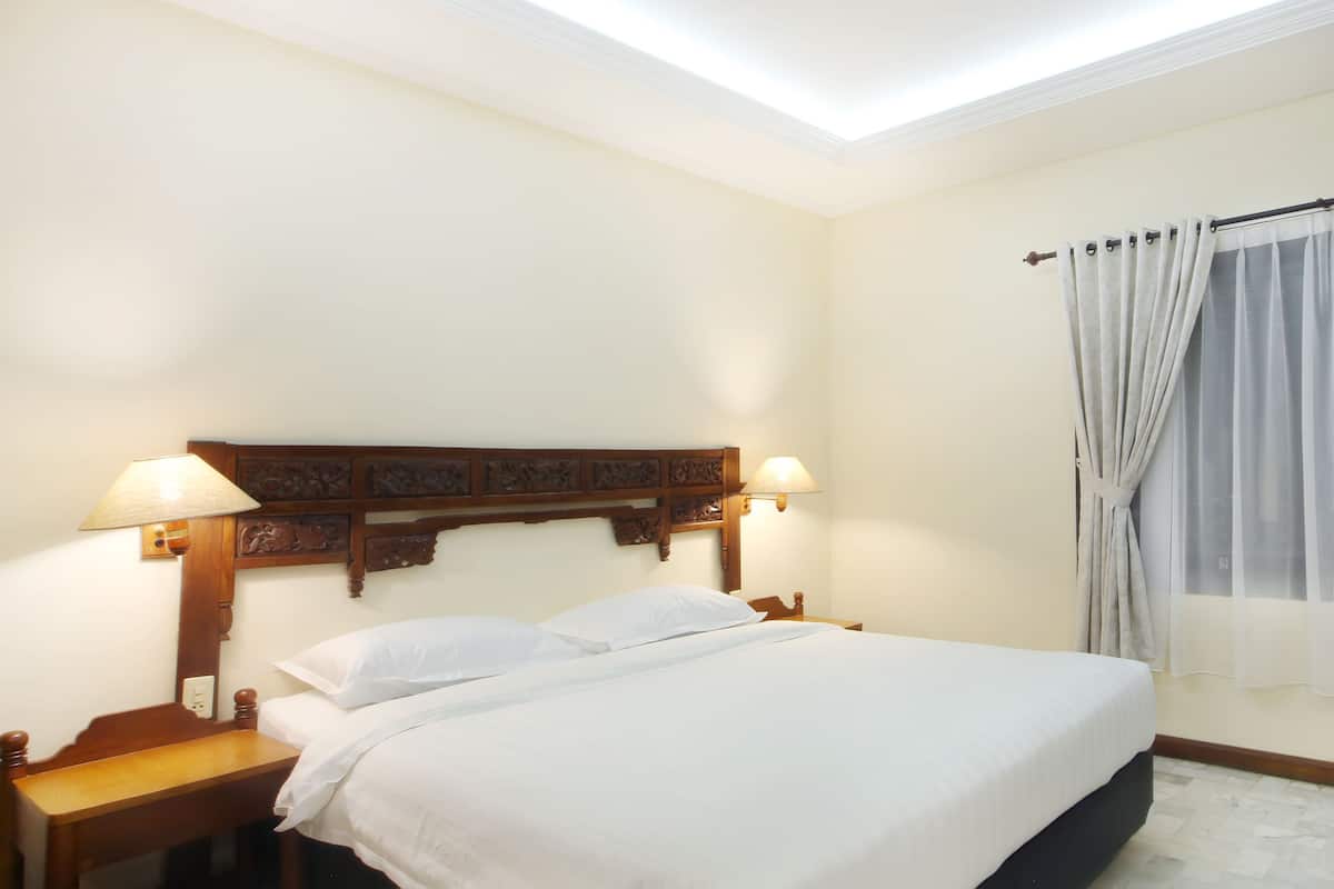 Quarto de hotel com uma cama grande, cabeceira de madeira, duas mesas de cabeceira com abajures e uma janela com cortinas.