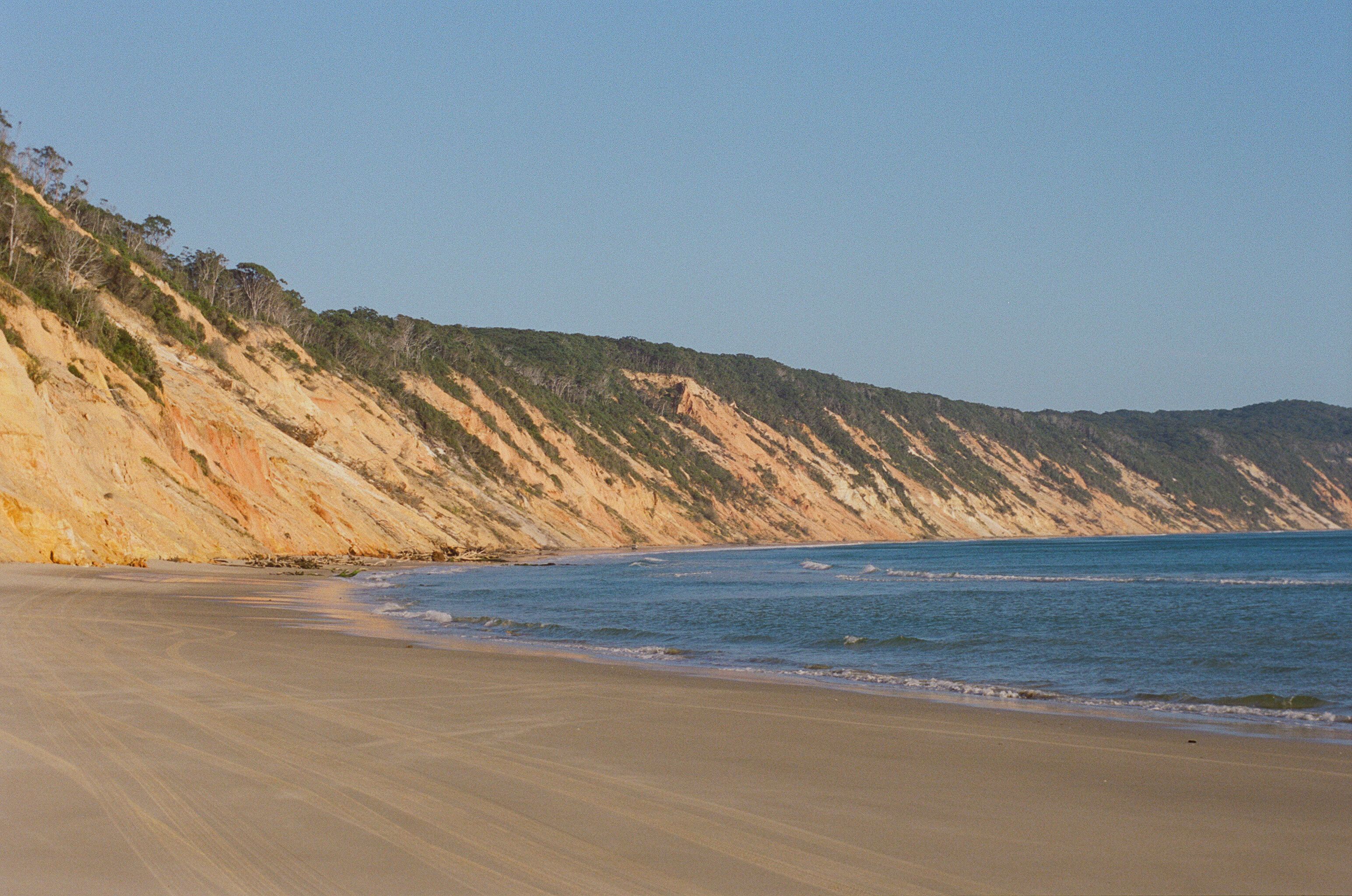 Uma praia com areal e um penhasco íngreme com vegetação esparsa.