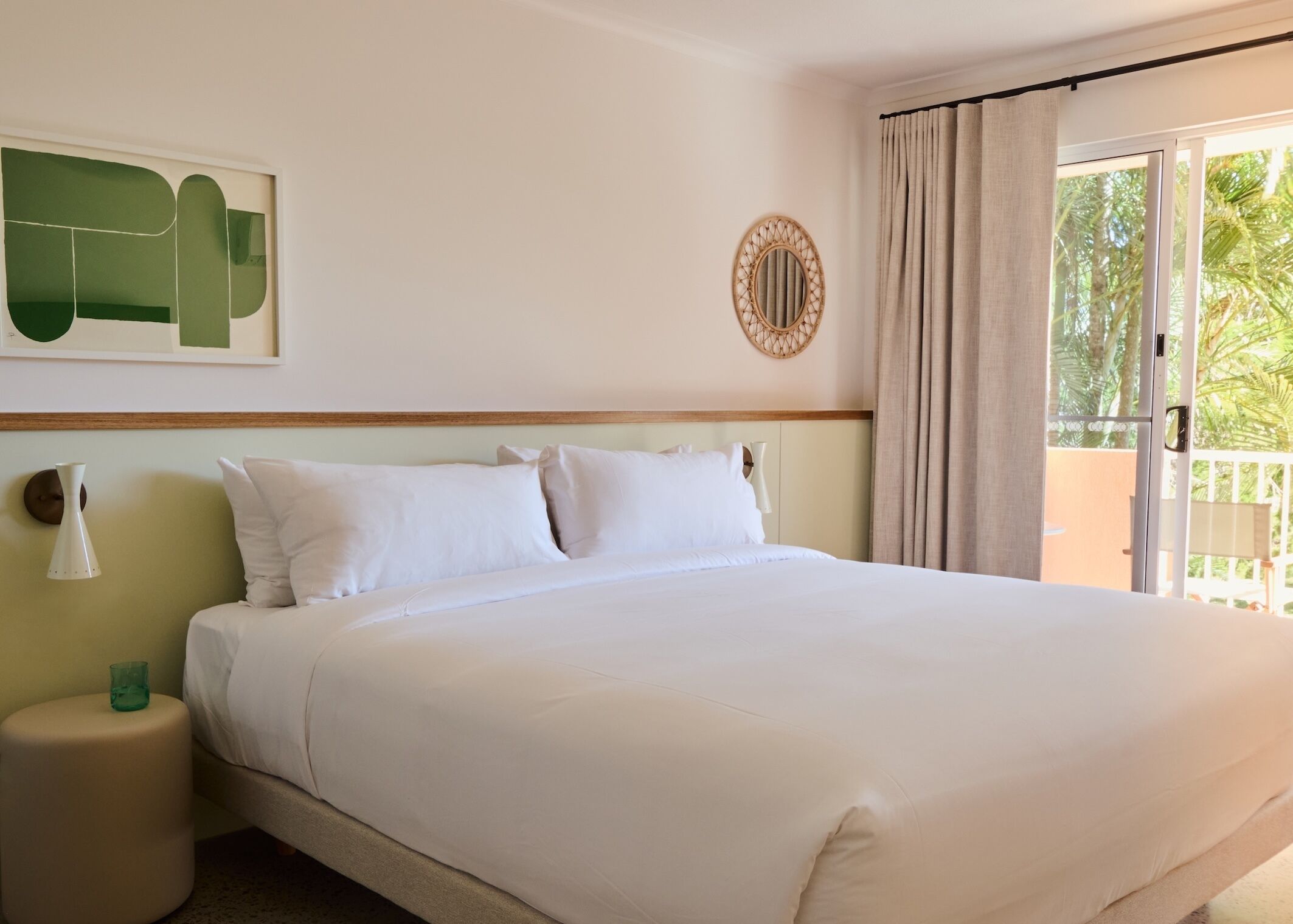 Quarto de hotel com uma cama grande, uma mesa de cabeceira, uma luminária, um espelho fixado na parede e vista para o verde pela janela.