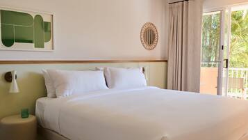 Quarto de hotel com uma cama grande, uma mesa de cabeceira, uma luminária, um espelho fixado na parede e vista para o verde pela janela.