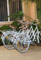 Uma fileira de bicicletas brancas estacionadas em frente a um edifício com grandes folhas verdes e uma janela ao fundo.