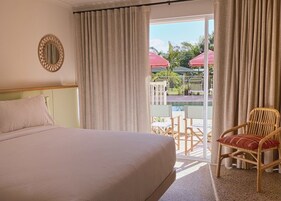 Quarto de hotel com cama, uma cadeira, janela com cortinas e vista para área externa com guarda-sóis.