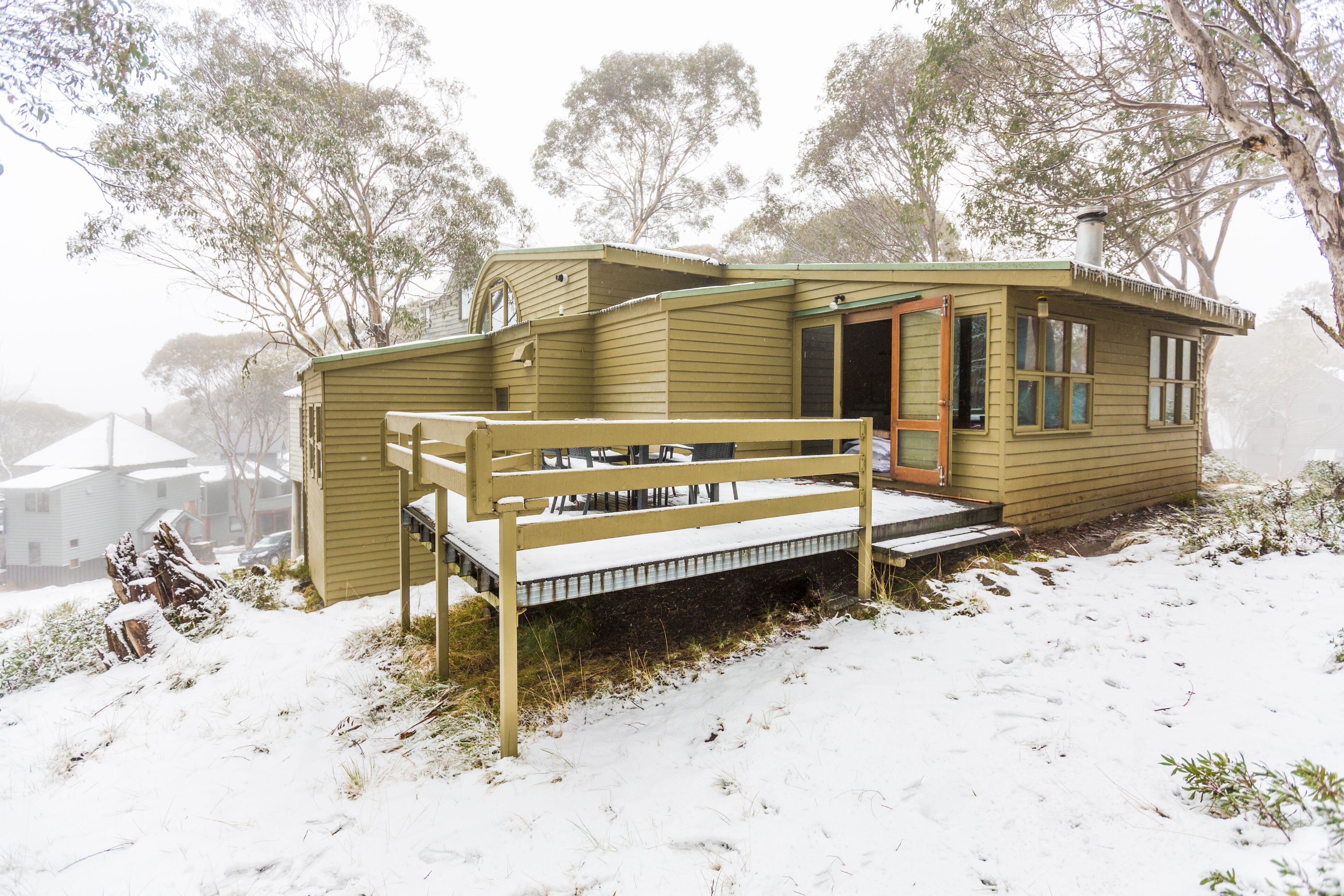 Uma casa pequena e de um único andar com varanda e porta, rodeada por neve e árvores.
