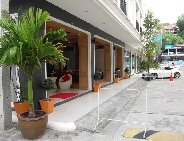 Entrada de um hotel com tapete vermelho, plantas em vasos e um carro branco estacionado na frente.