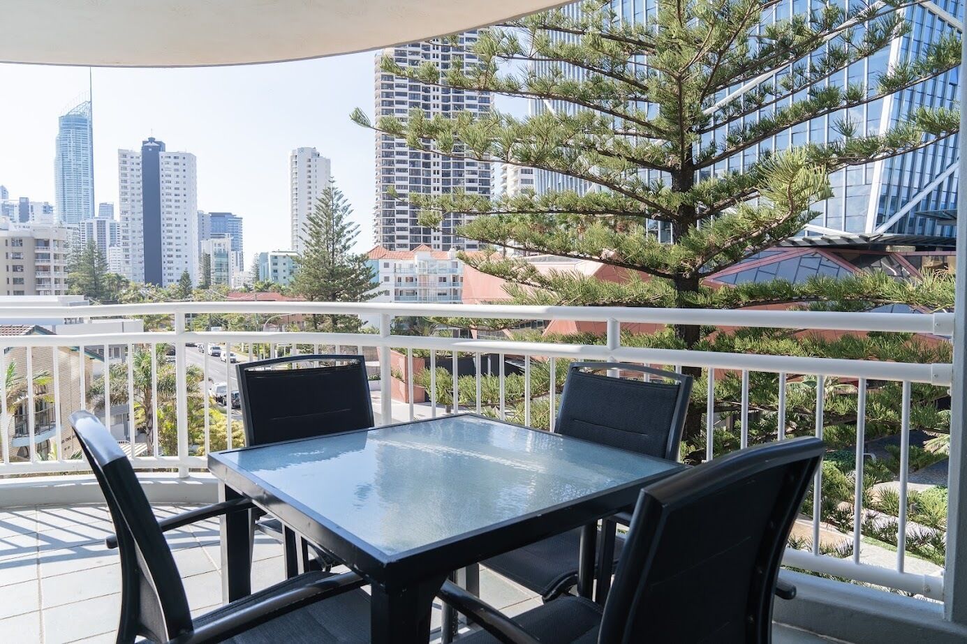 Uma varanda com mesa e cadeiras de vidro, com vista para um horizonte urbano com edifícios modernos e uma árvore grande.