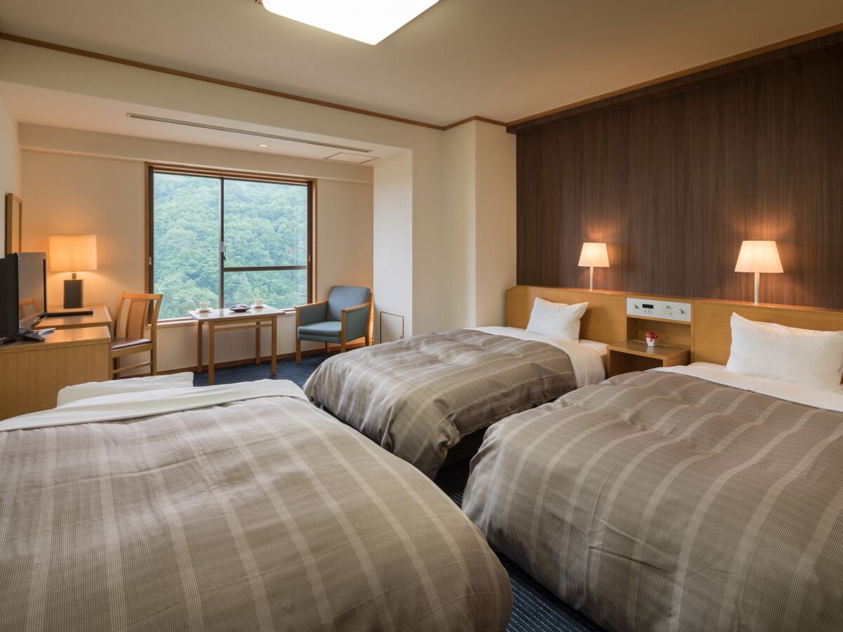 Quarto de hotel com duas camas, uma escrivaninha com cadeira, televisão e janela com vista para árvores.