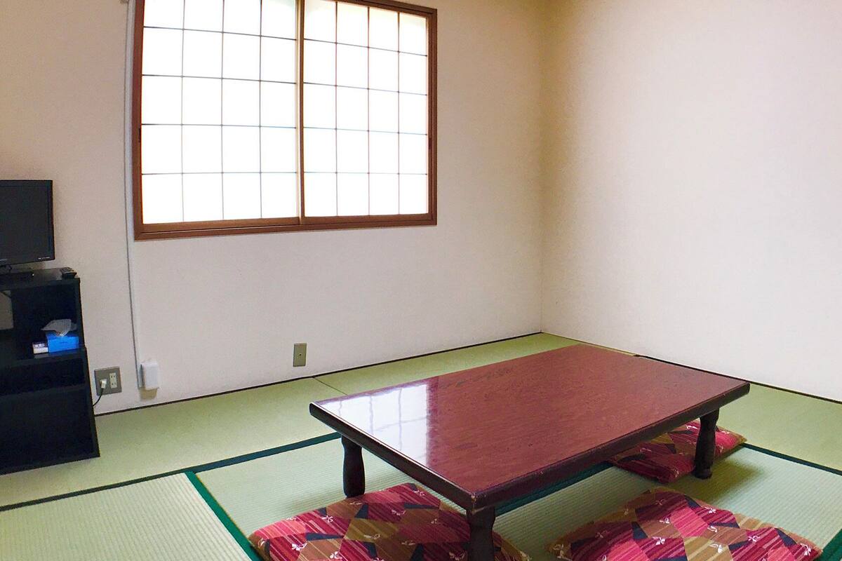 Quarto com esteiras de tatame, uma mesa baixa, uma televisão numa prateleira e uma janela com vidros em grade.
