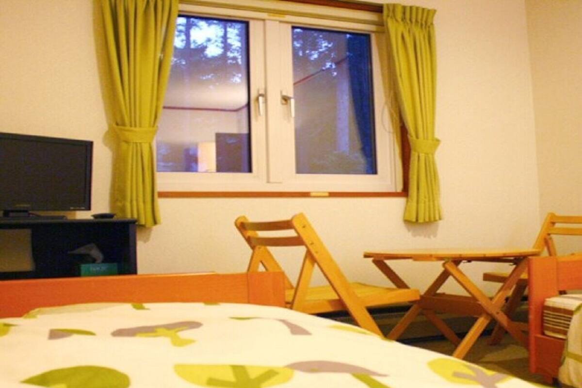 Quarto de hotel com uma cama, televisão, janela com cortinas e duas cadeiras de madeira.