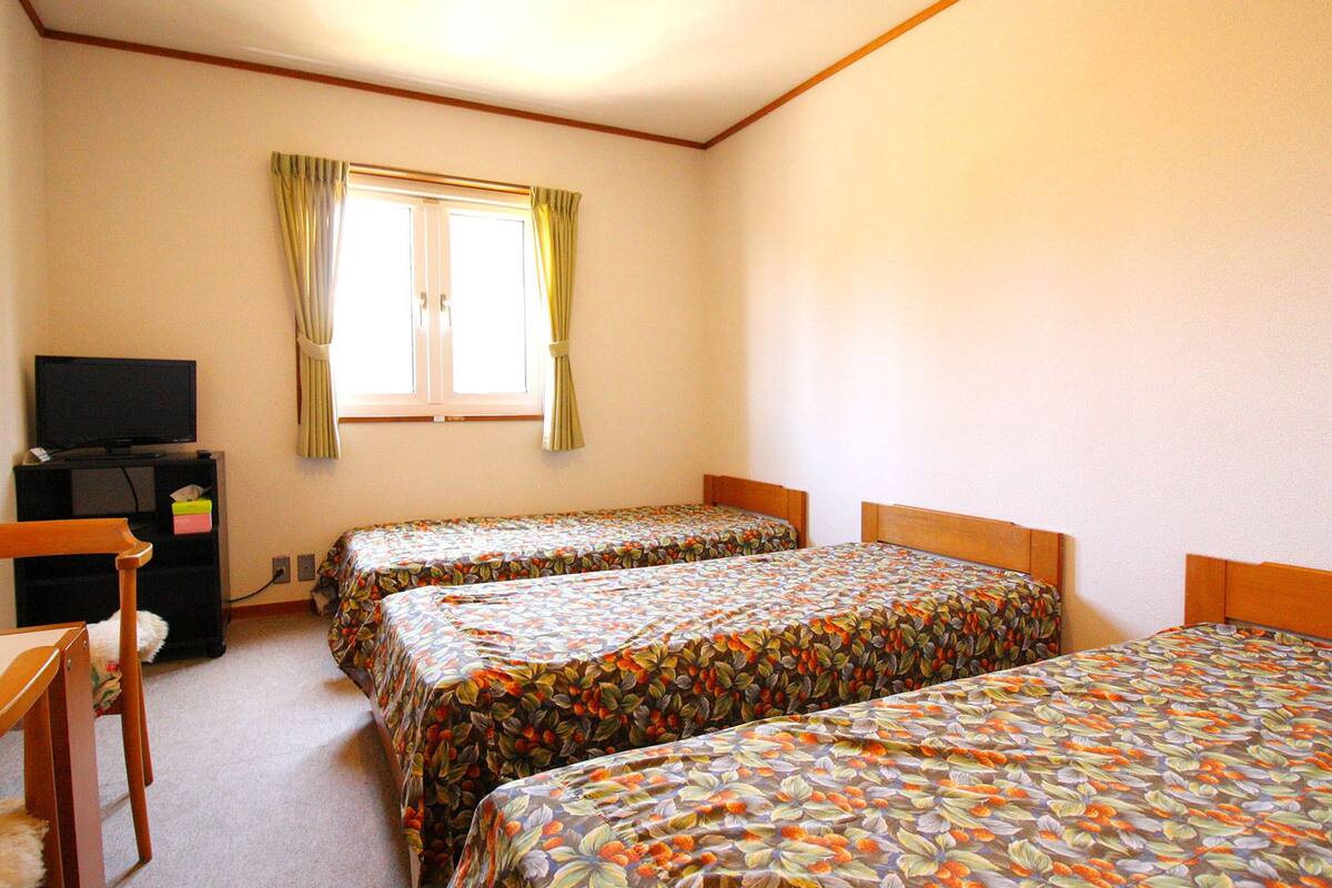 Quarto com duas camas, uma escrivaninha, televisão e janela com cortinas.