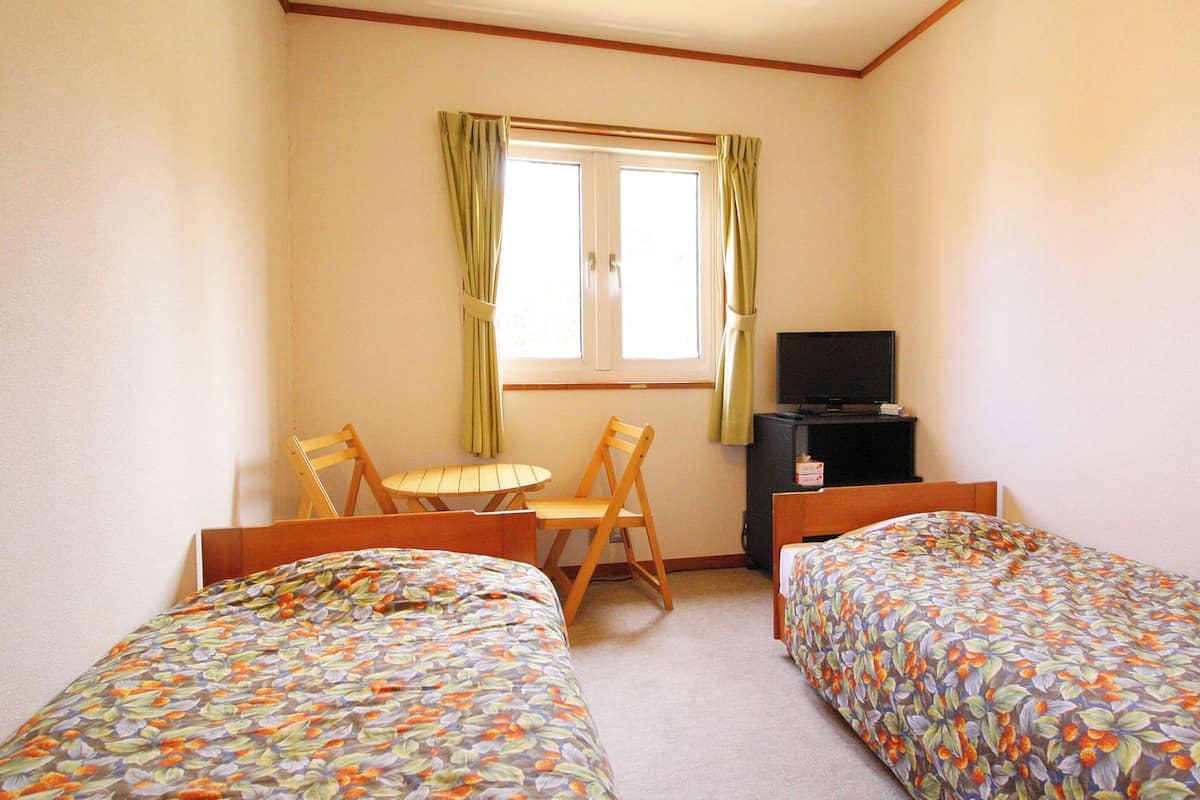 Quarto com duas camas, uma mesa pequena, uma cadeira, televisão e janela com cortinas.