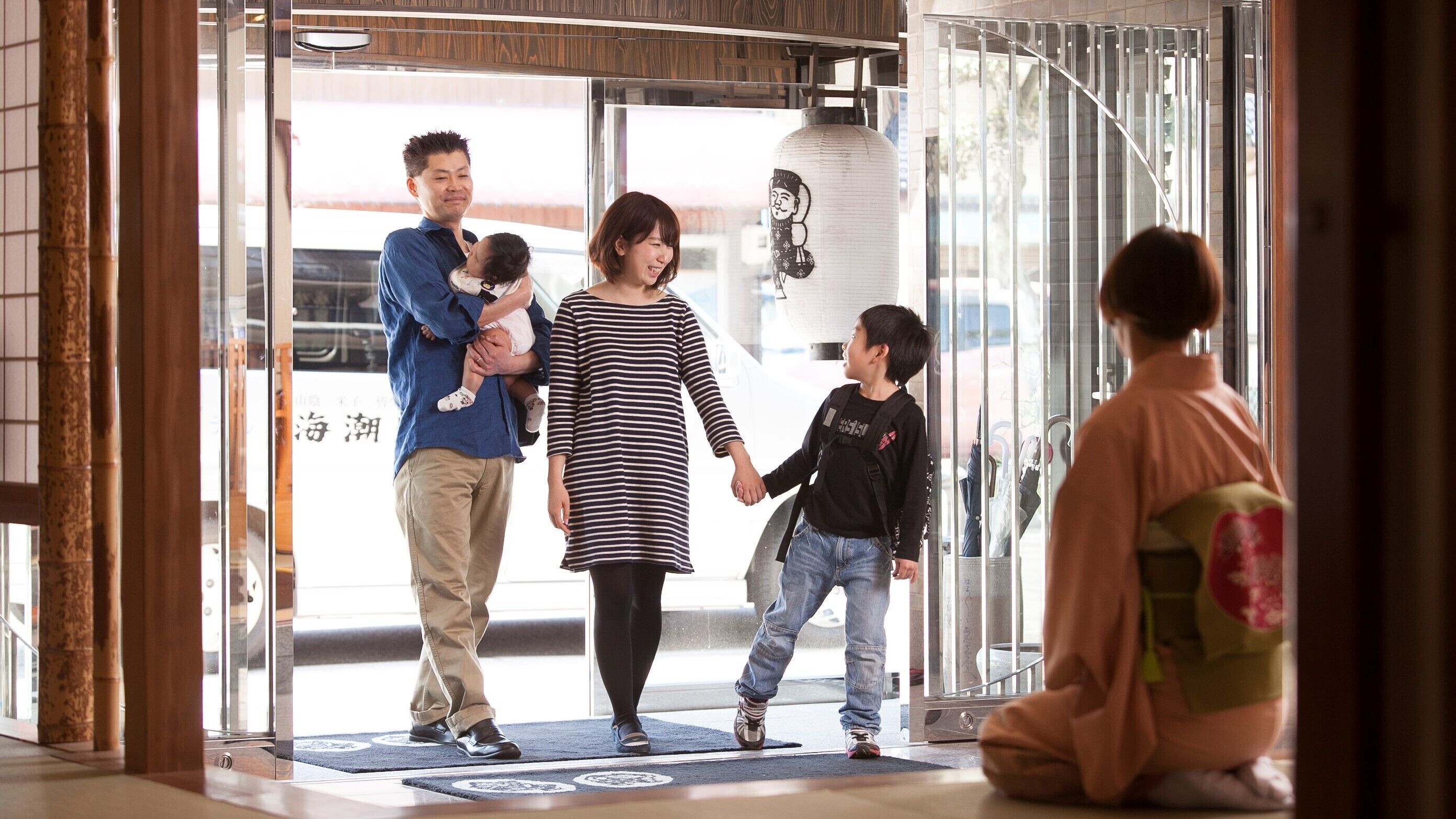 Uma família entrando em um prédio por uma porta de vidro, com uma criança de mãos dadas com um adulto.