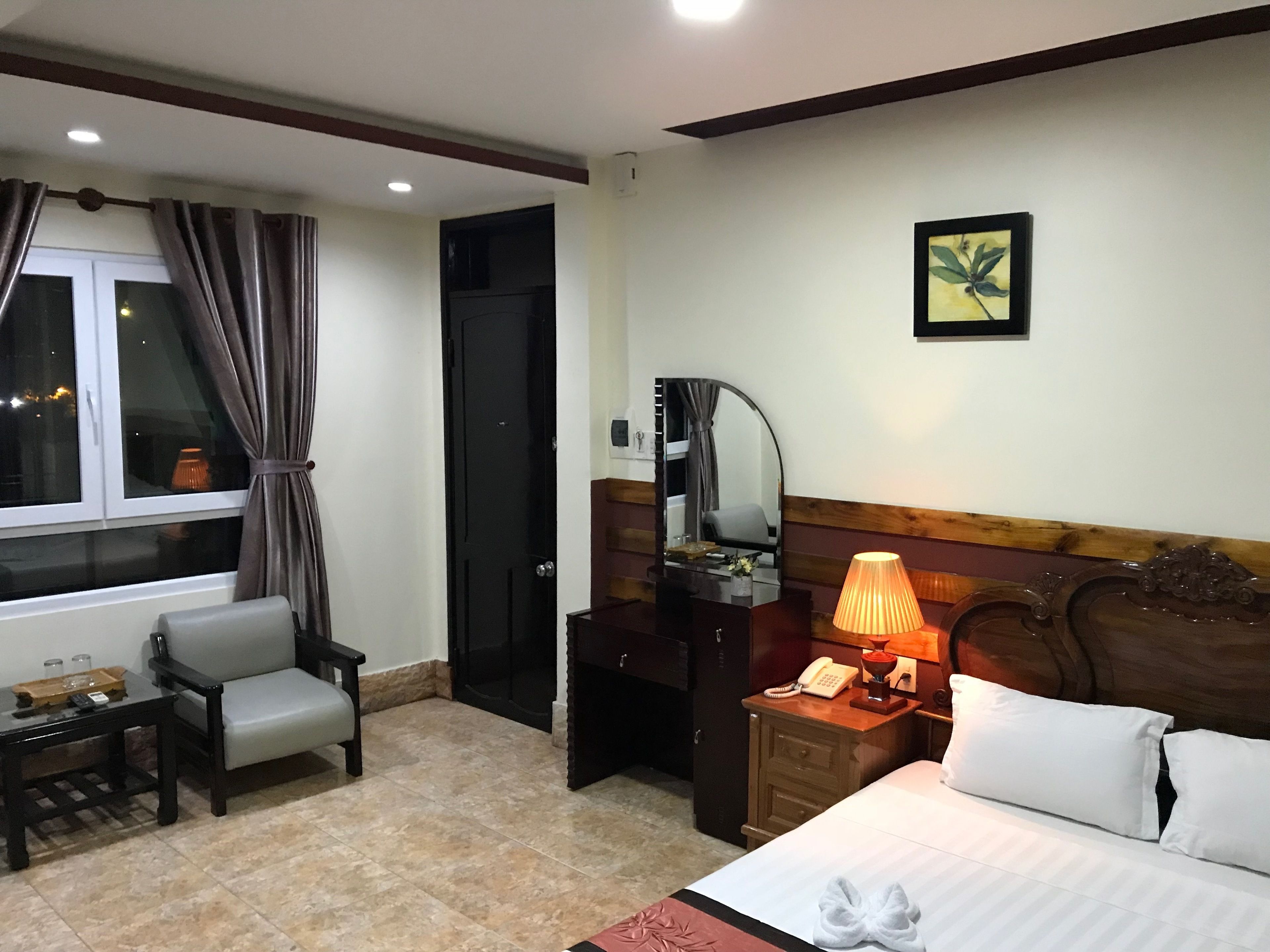 Quarto de hotel com cama, escrivaninha com luminária, cadeira, cômoda e janela com cortinas.