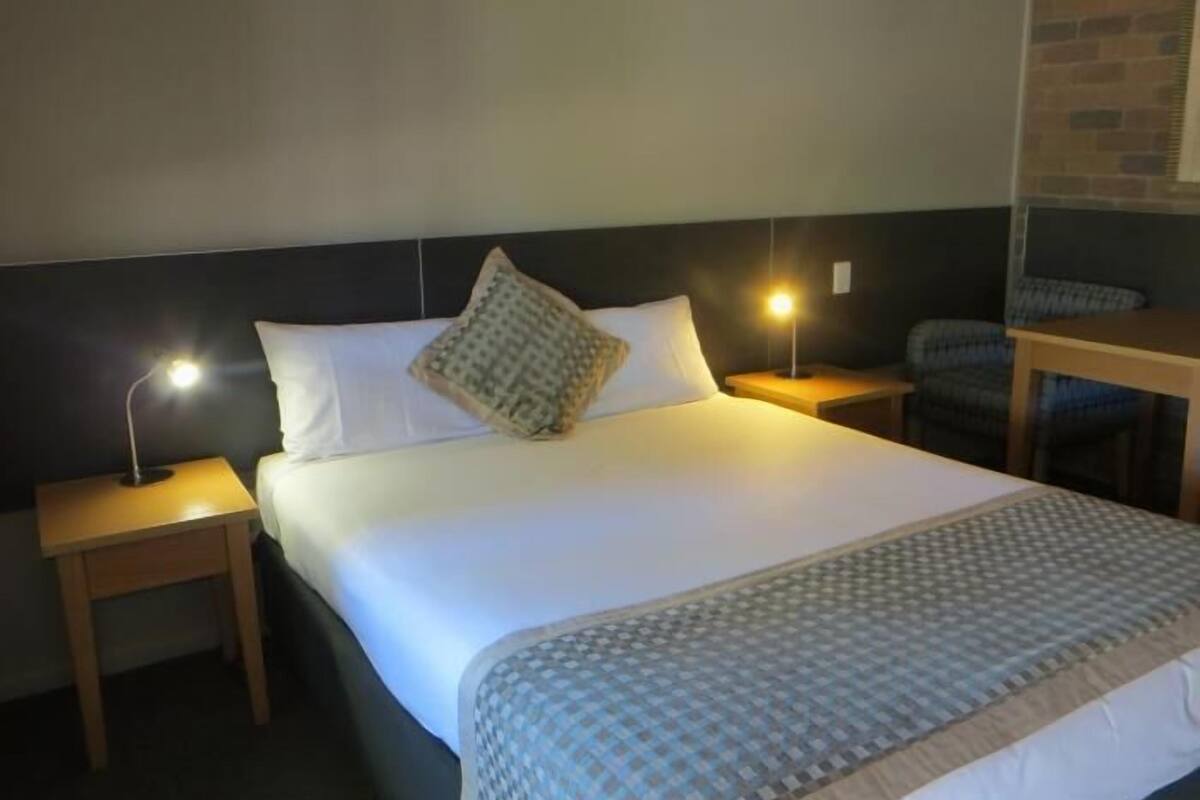 Cama bem arrumada com colcha estampada, duas luminárias de cabeceira e uma cadeira em quarto com paredes escuras e parede de tijolos decorativa.