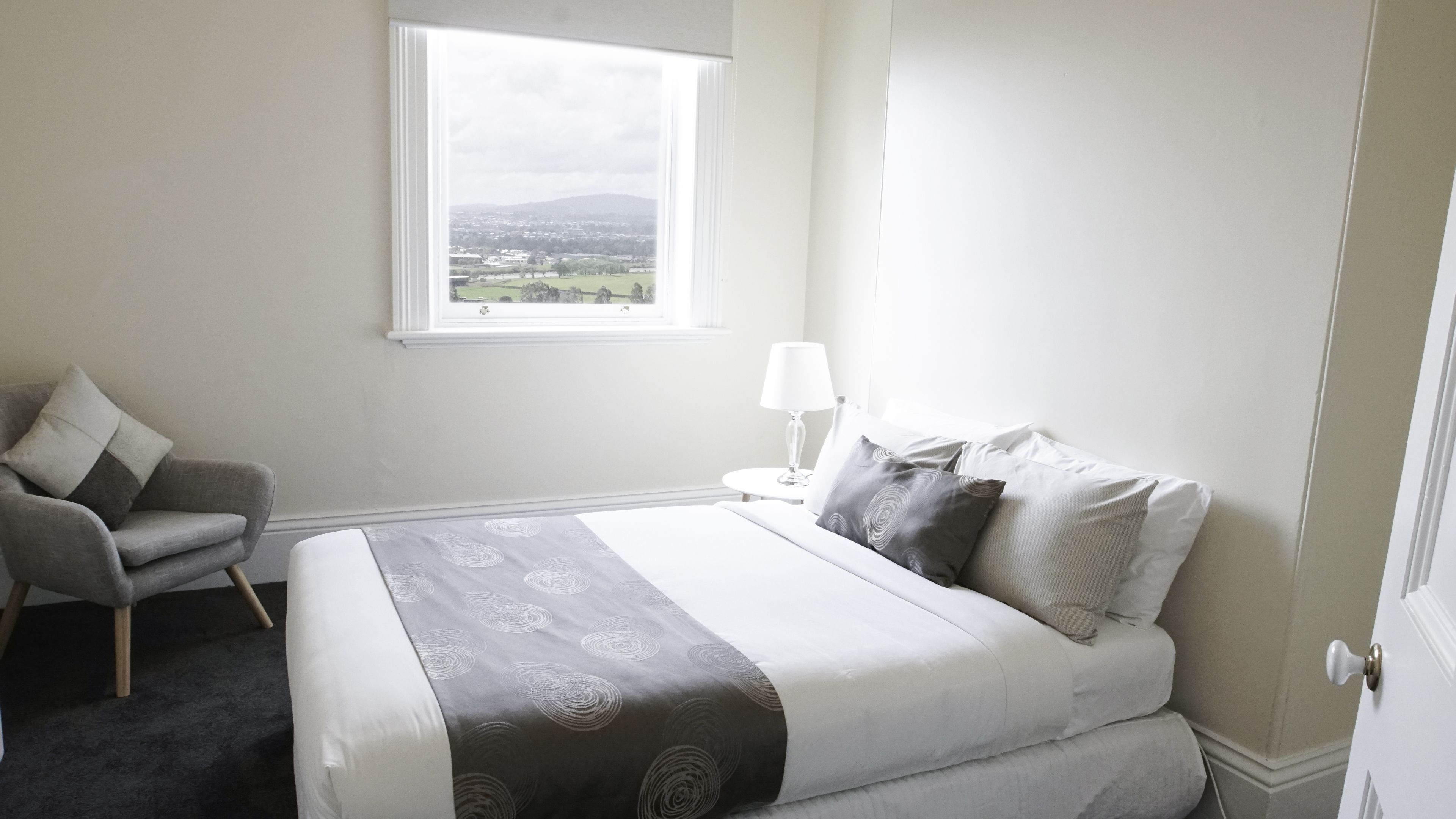 Quarto com cama, uma cadeira, mesa de cabeceira, luminária e janela com vista para a paisagem.