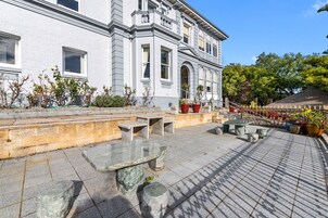 Um pátio com bancos de pedra, um edifício com janelas em arco e plantas em vasos.