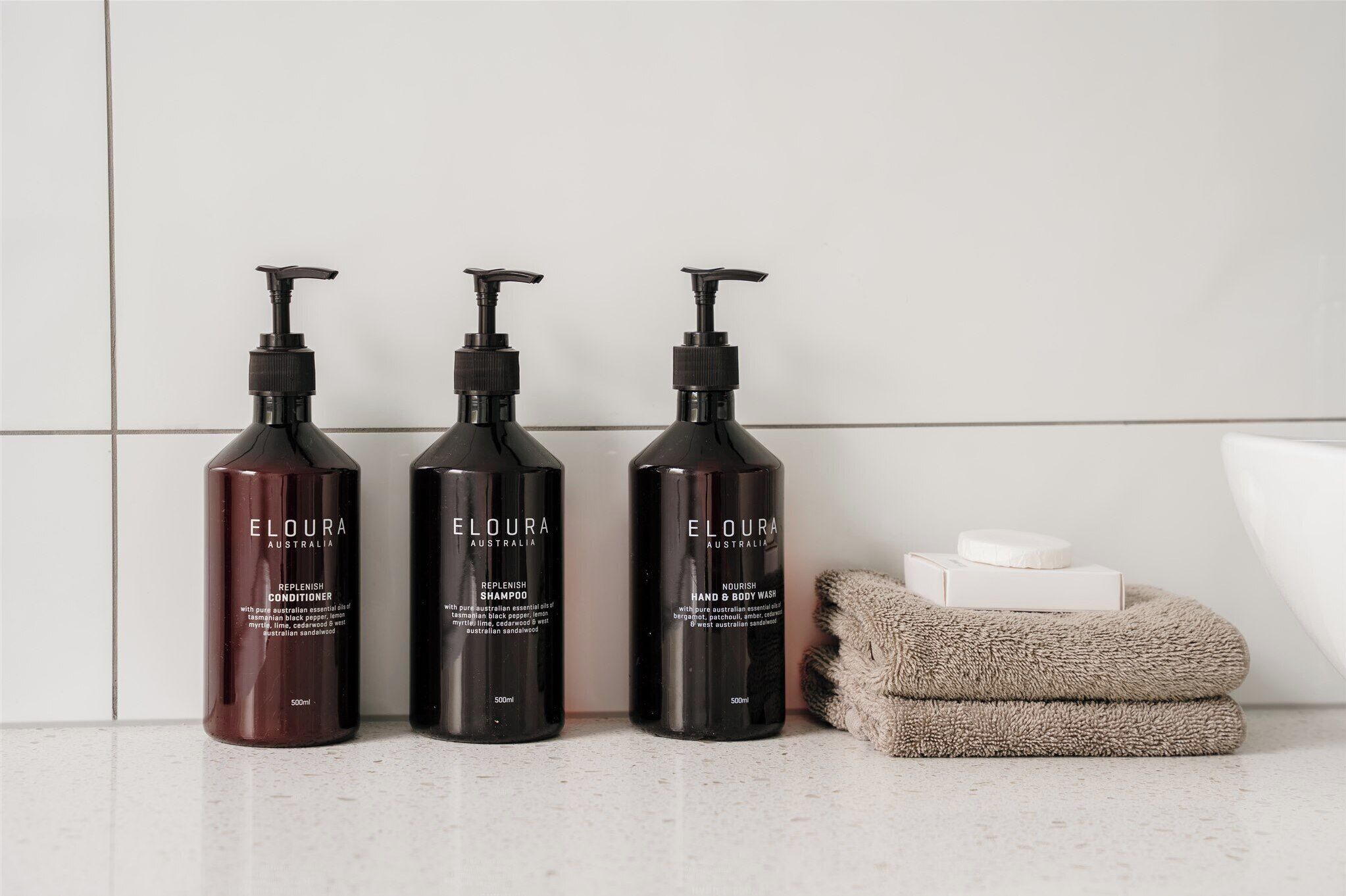 Três frascos de produtos de cuidados para a pele Eloura Australia e uma toalha dobrada em uma bancada de banheiro.