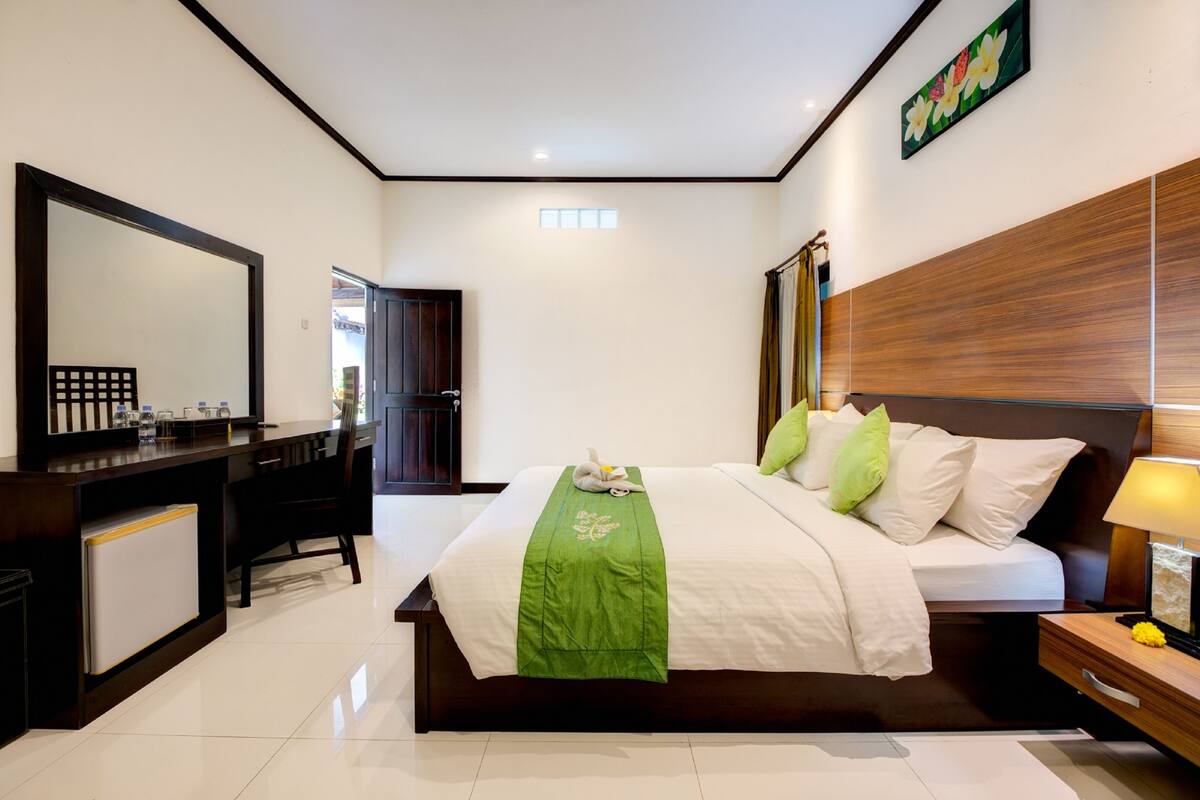 Quarto de hotel com cama, mesa de cabeceira, abajur e escrivaninha com espelho.