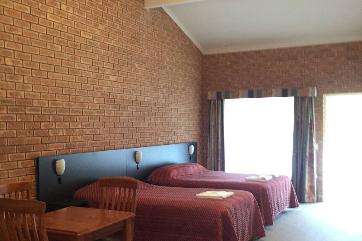 Quarto de hotel com duas camas, uma parede de tijolos, uma luminária redonda no teto e vista para o exterior.
