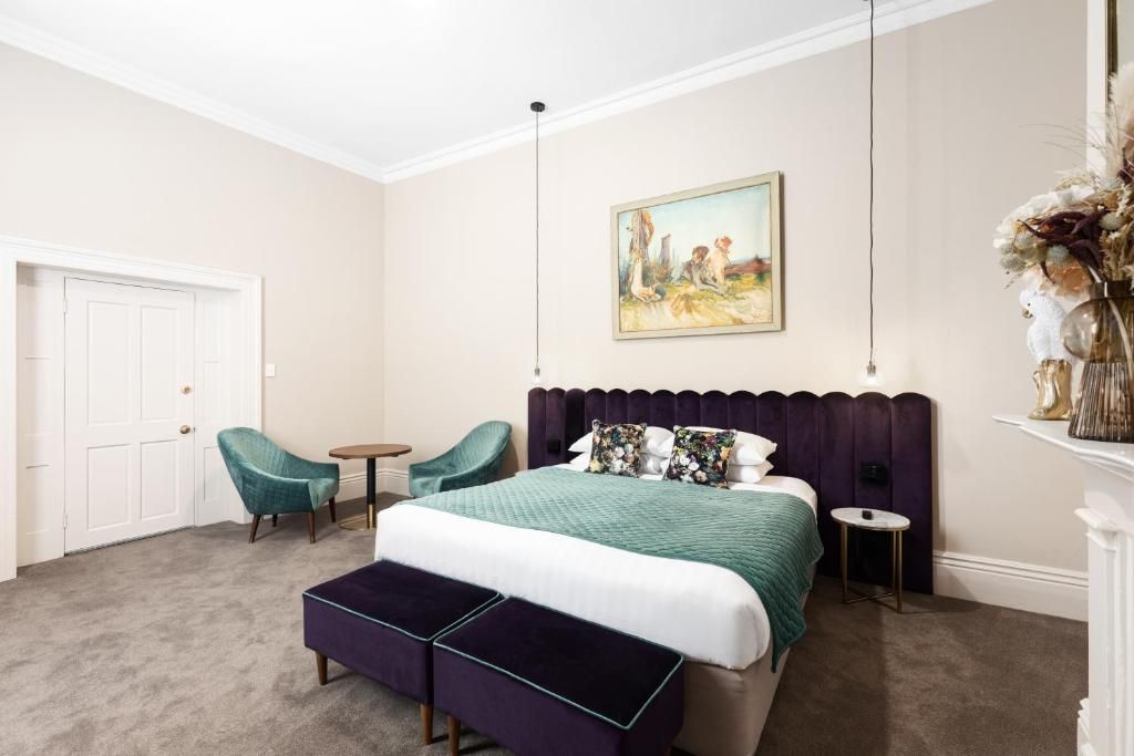 Quarto com cama, duas cadeiras, uma mesinha, um banco e um quadro na parede.