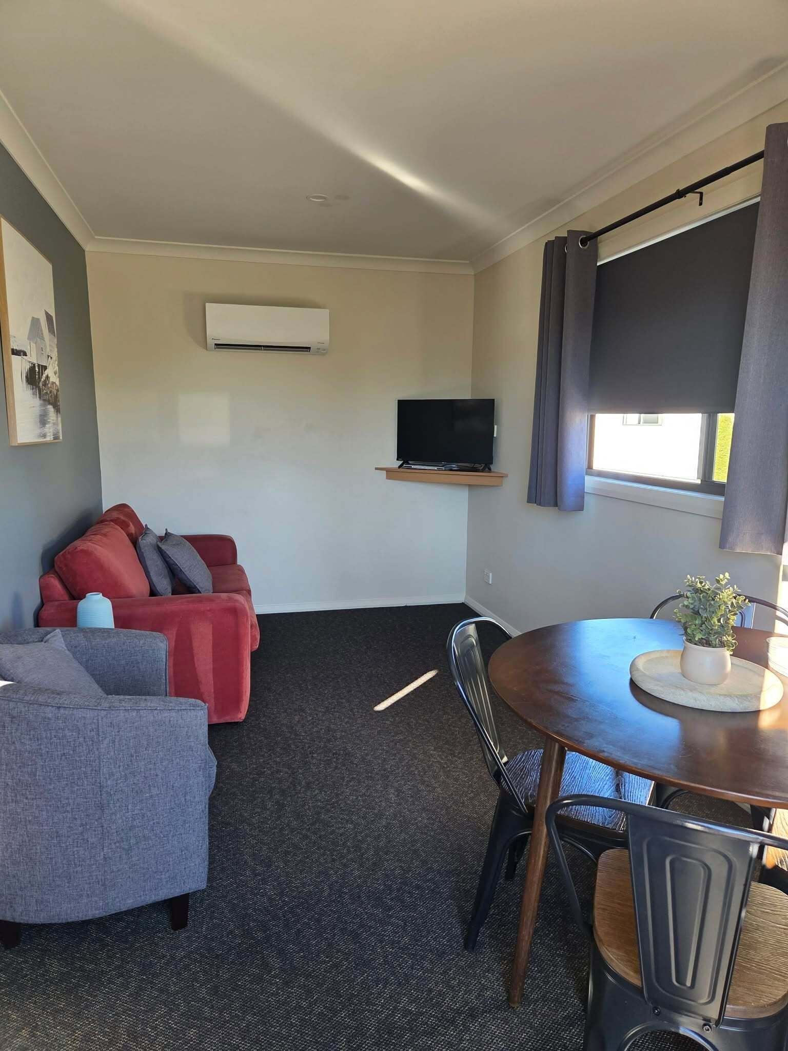Quarto com sofá vermelho, poltrona cinza, mesa de jantar de madeira com cadeiras, televisão de tela plana na parede e janela com cortinas.