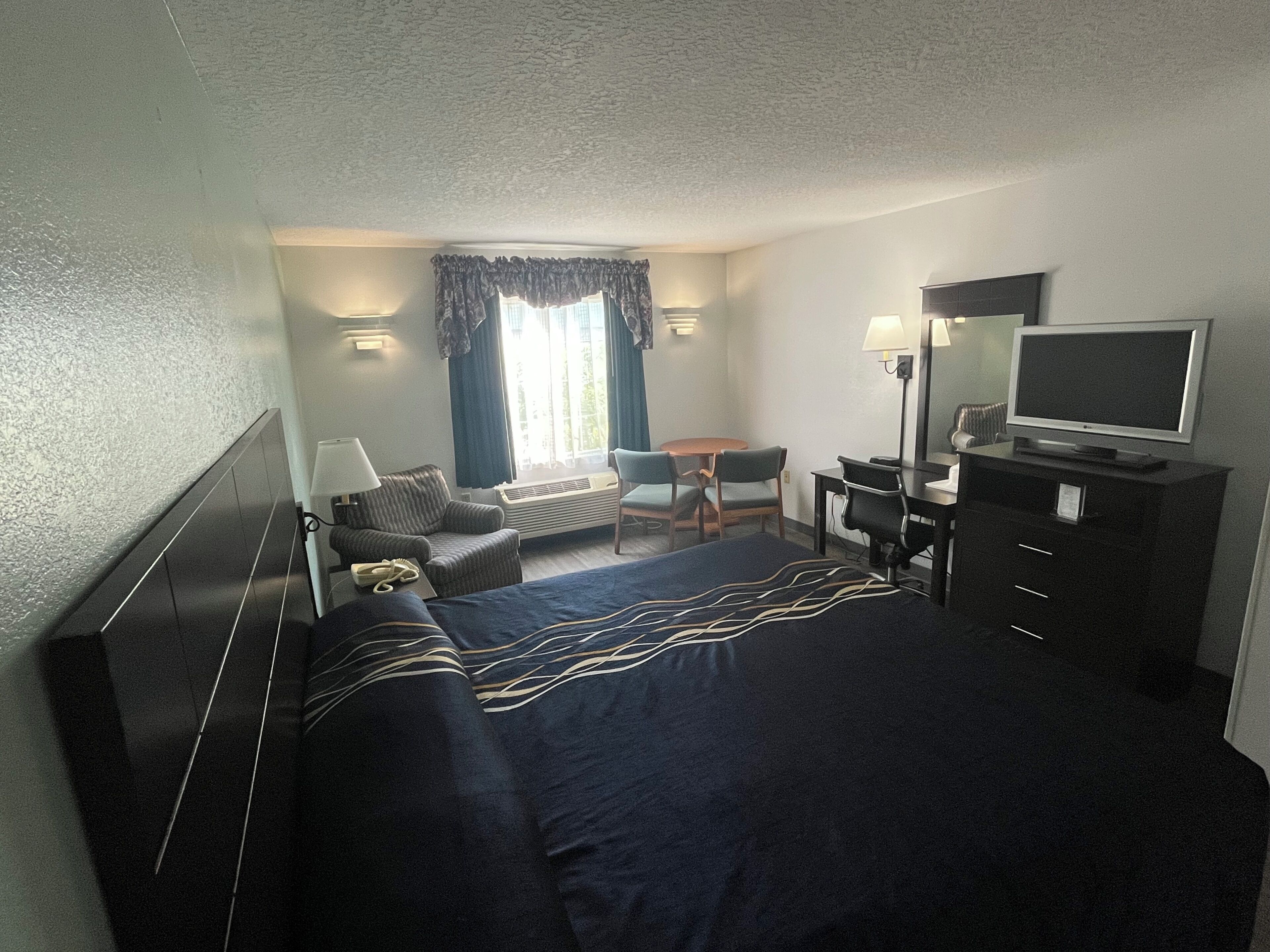 Quarto de hotel com cama, escrivaninha, cadeira, televisão e janela com cortinas.