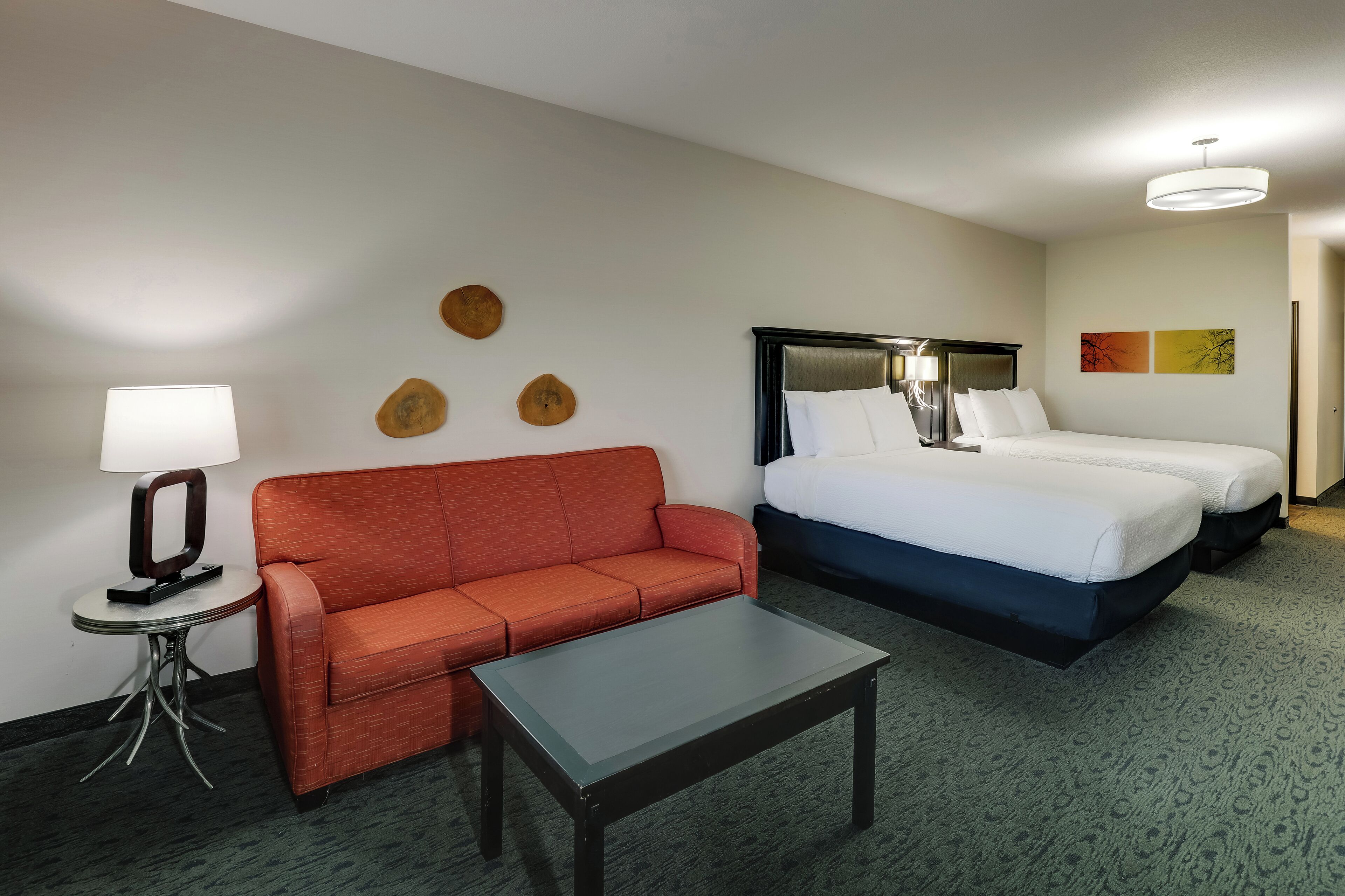 Quarto de hotel com sofá vermelho, uma mesinha, uma cama com dois travesseiros e uma luminária.