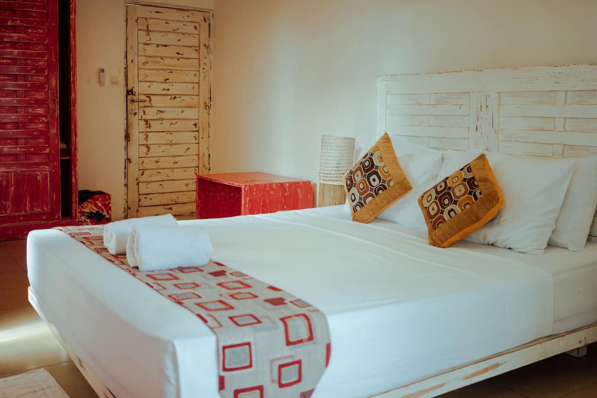 Uma cama com roupa de cama branca e almofadas estampadas, um criado-mudo vermelho e uma porta de madeira ao fundo.