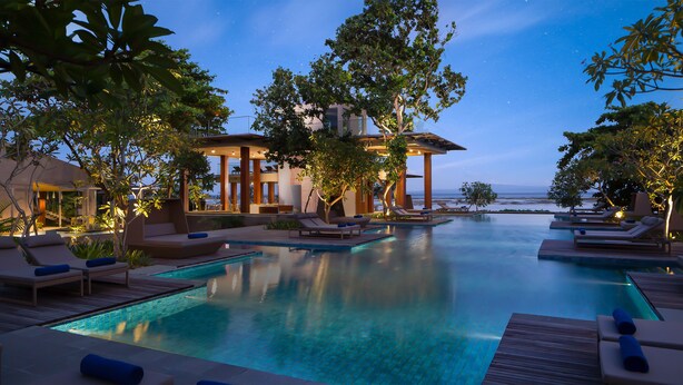 Uma área de piscina tranquila com espreguiçadeiras e um gazebo, cercada por árvores e com vista para um corpo d'água.