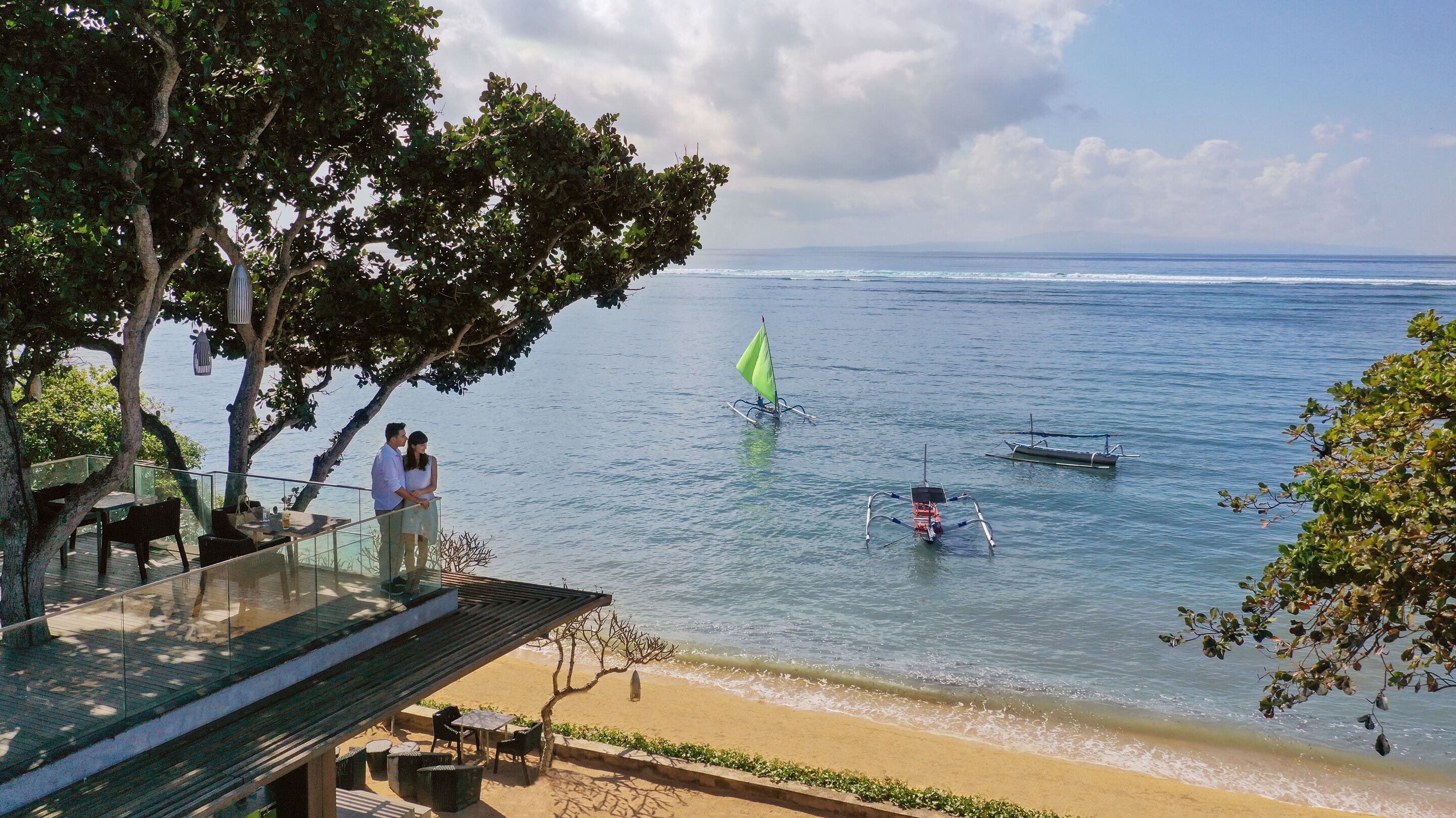 Vista à beira-mar com guarda-corpo de vidro, um casal em um deck e um veleiro na água.