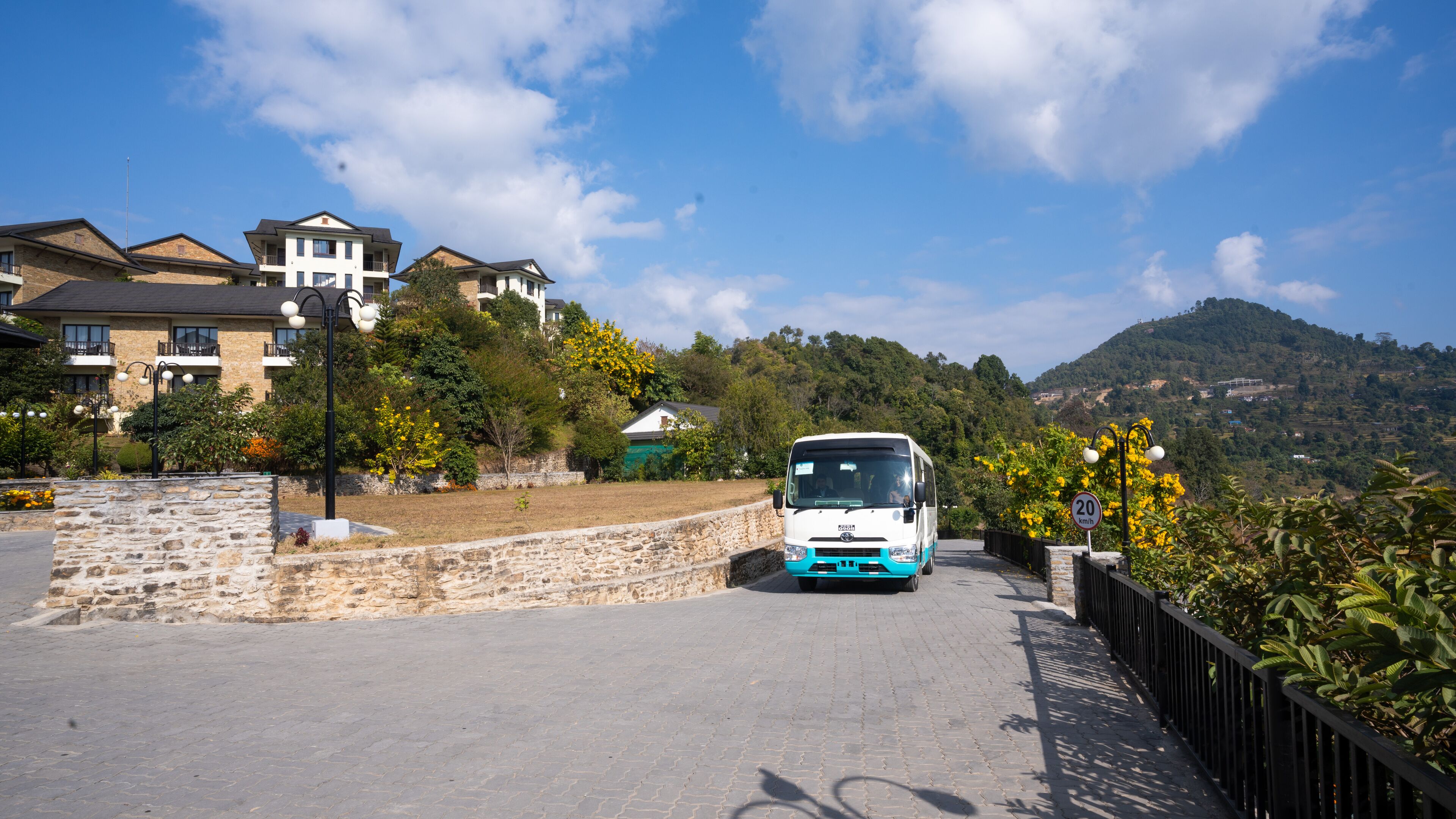 Um ônibus em uma estrada pavimentada, com paredes de pedra e vegetação em um lado, e uma paisagem montanhosa ao fundo.