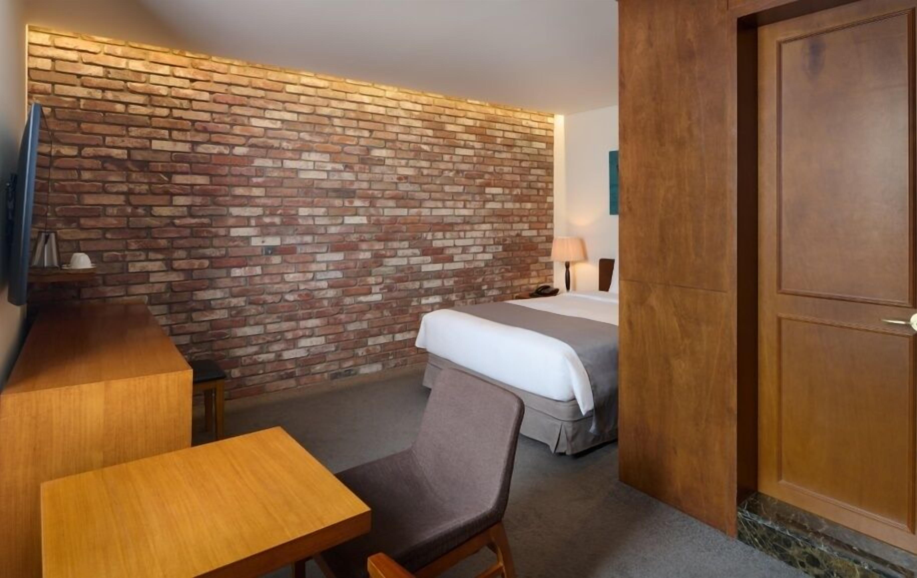 Quarto de hotel com parede de tijolos, uma cama, uma escrivaninha e uma cadeira.