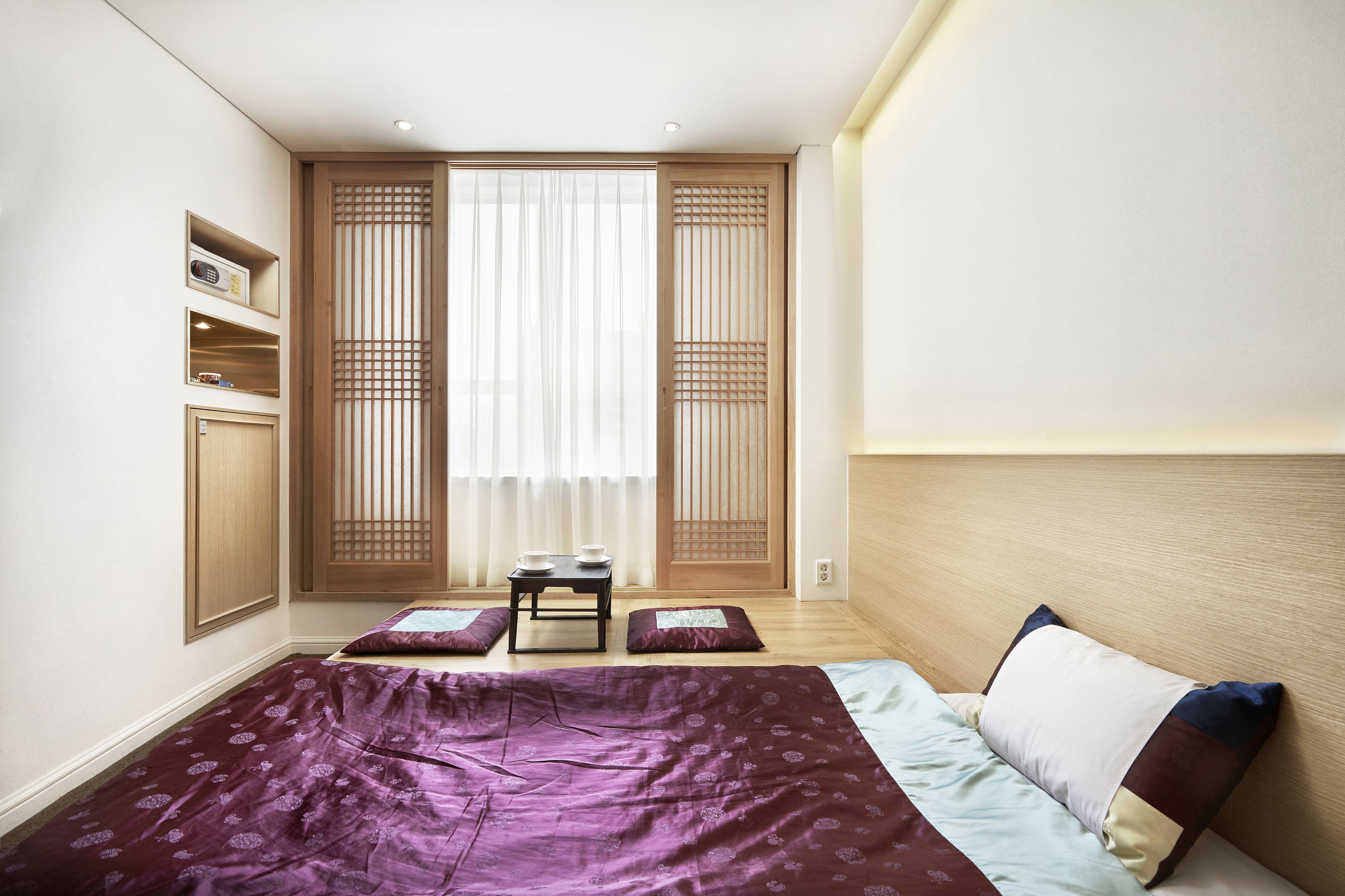 Quarto com cama, almofadas, uma mesinha e janela com persianas.